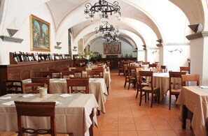 Um restaurante com várias mesas e cadeiras, um candelabro e pinturas nas paredes.