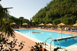 Piscina com espreguiçadeiras e guarda-sóis, cercada por árvores e colinas.