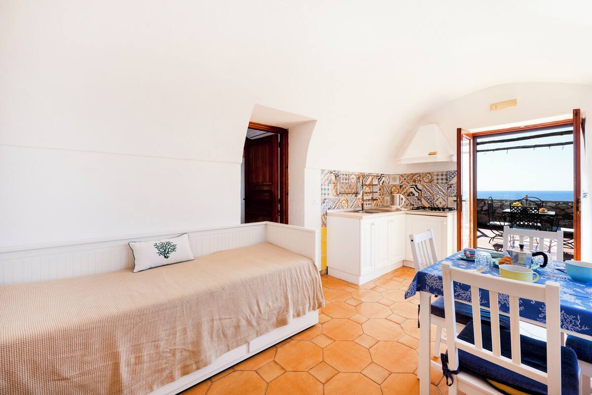 Quarto compacto com cama, mesa de jantar com cadeiras, área de cozinha e vista para o mar.