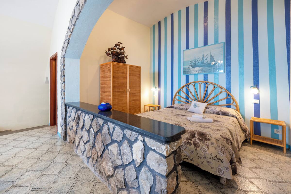 Um quarto com bancada de pedra, cama com colcha florida, armário de madeira e parede listrada com uma pintura.