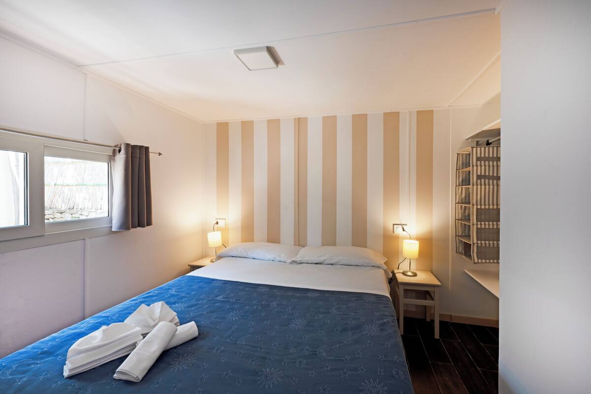 Quarto de hotel com cama, duas luminárias de cabeceira, janela com cortinas, colcha azul e uma pequena mesa de cabeceira.