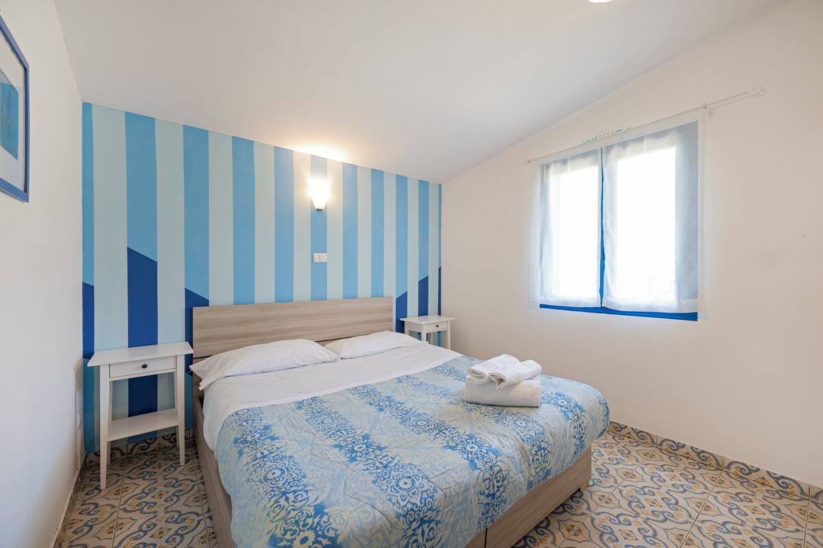 Quarto com cama, mesa de cabeceira, luminária na parede e janela com cortinas.