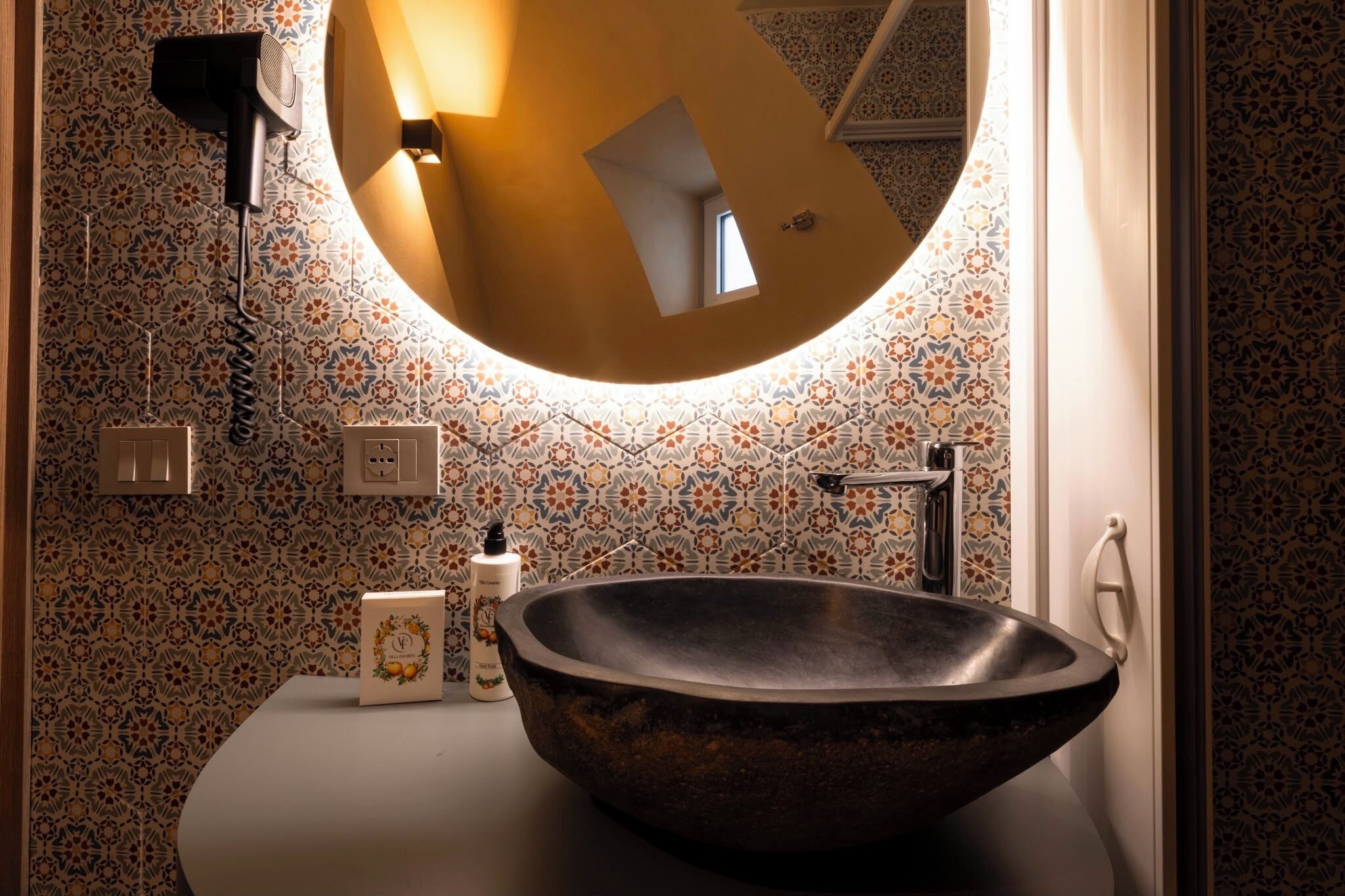 Casa de banho com espelho redondo, lavatório moderno e paredes com azulejos estampados.