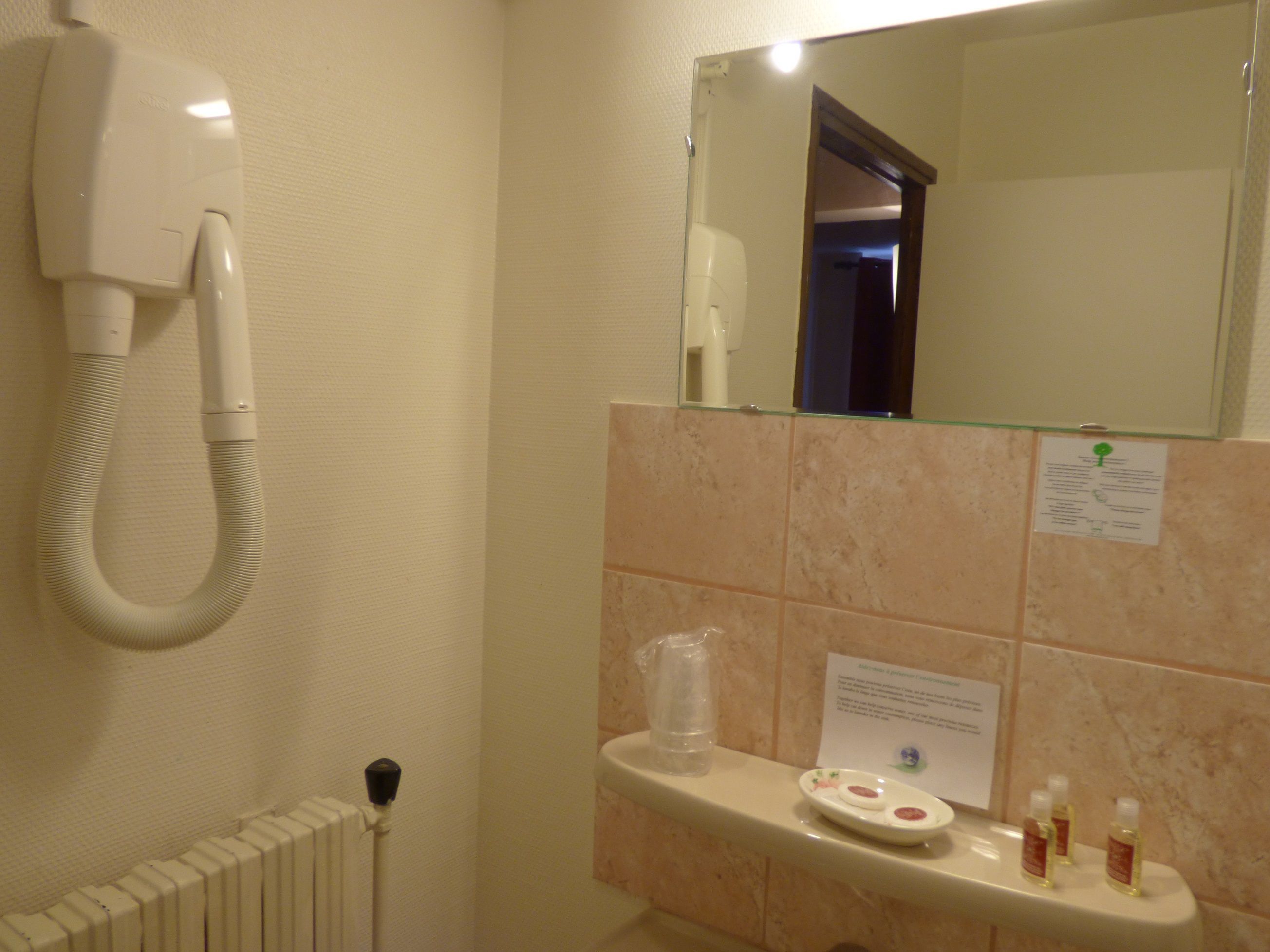 Casa de banho com secador de cabelo fixado na parede, radiador, espelho com prateleira de vidro e um aviso em papel.