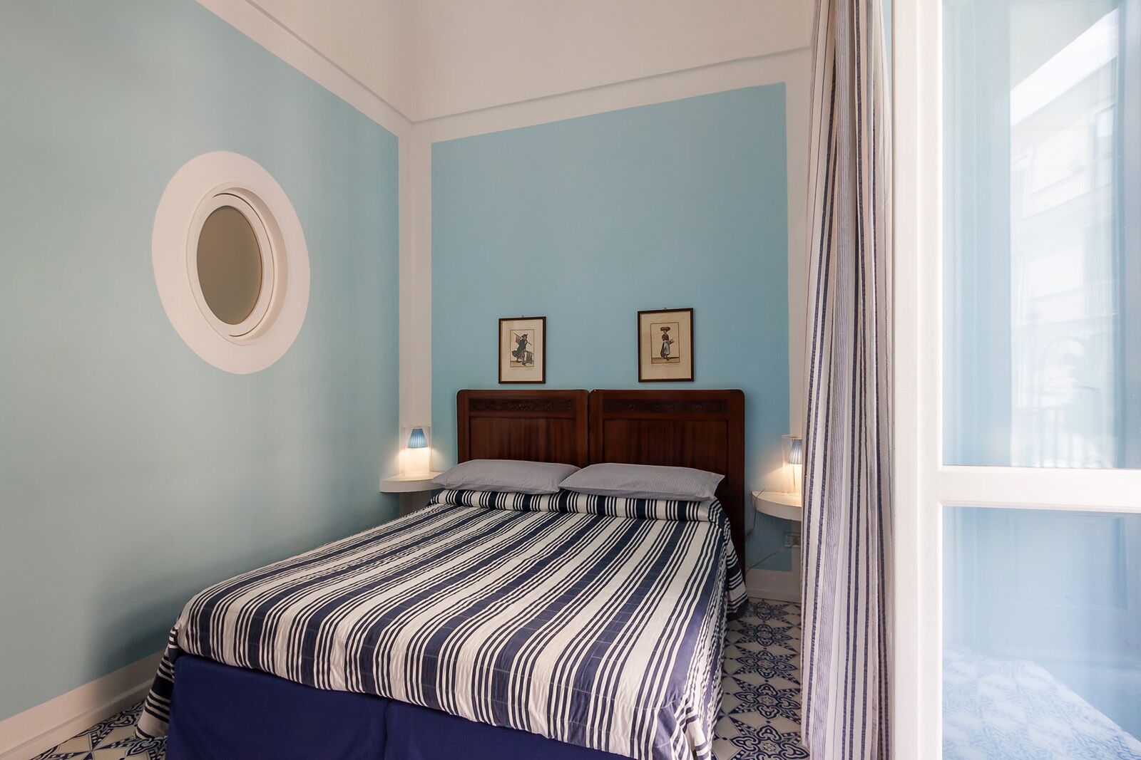 Quarto com uma cama, duas mesas de cabeceira, um espelho redondo e duas imagens emolduradas na parede.