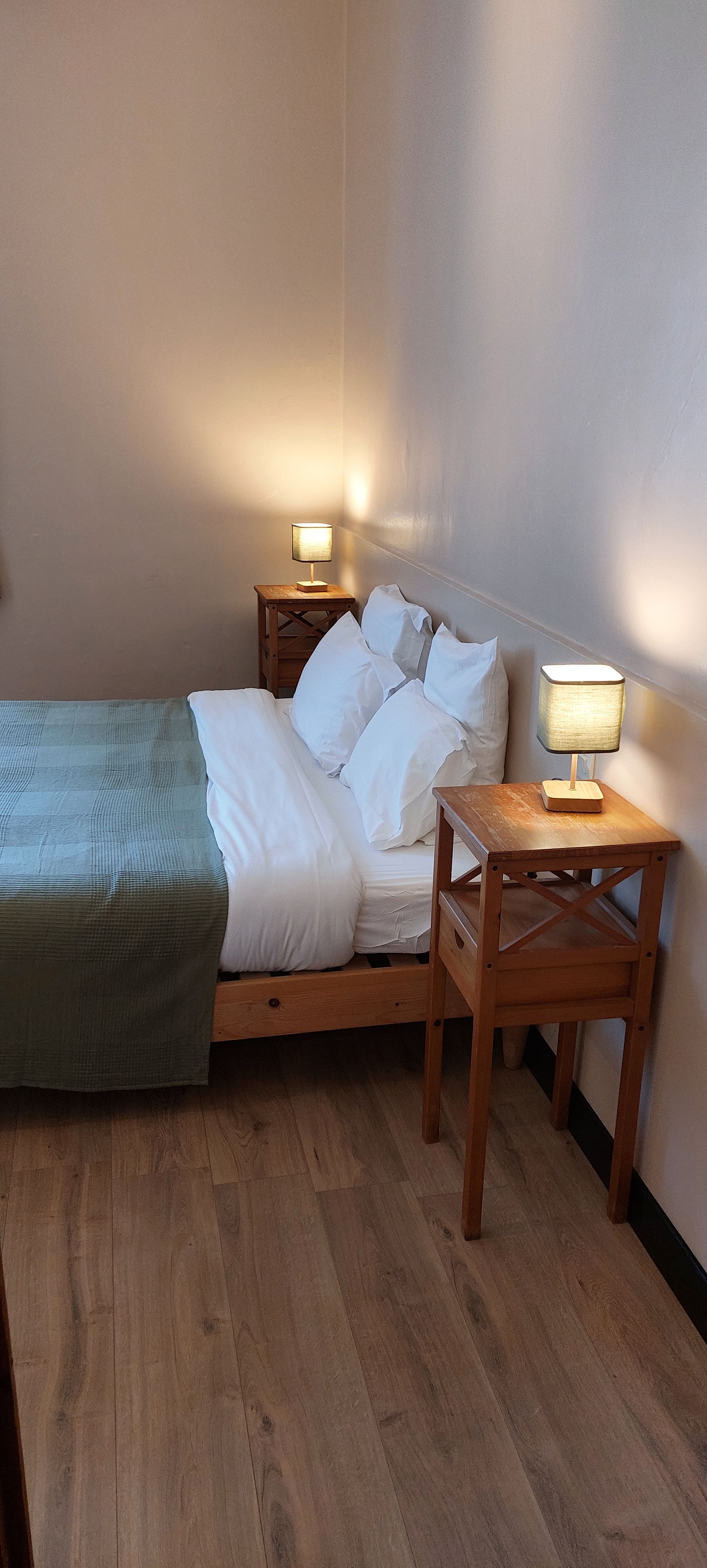 Uma cama de solteiro com estrutura de madeira, duas mesas de cabeceira com abajures e piso de madeira.