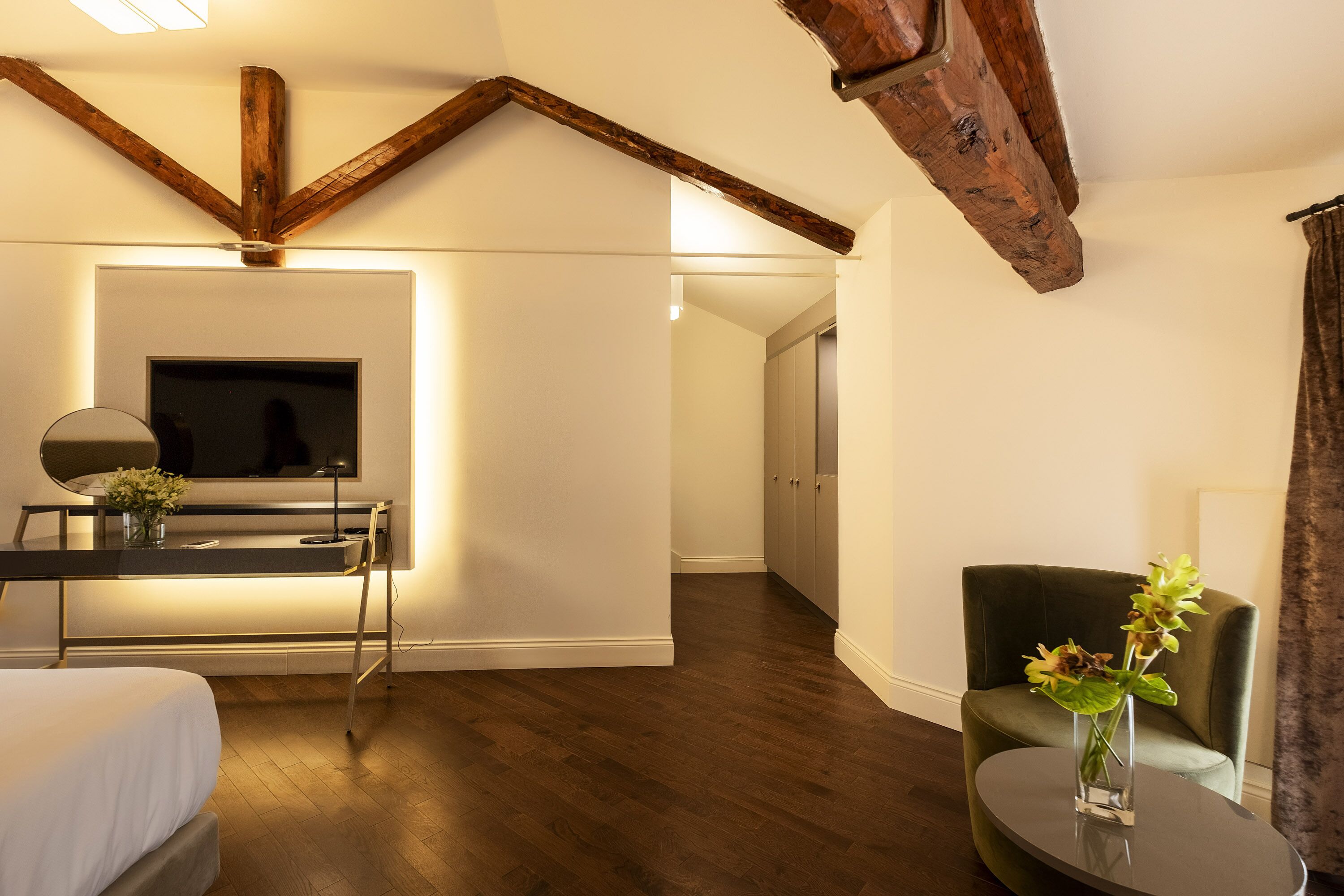 Quarto de hotel moderno com teto de madeira, cama, secretária com espelho, poltrona verde e um vaso com flores.