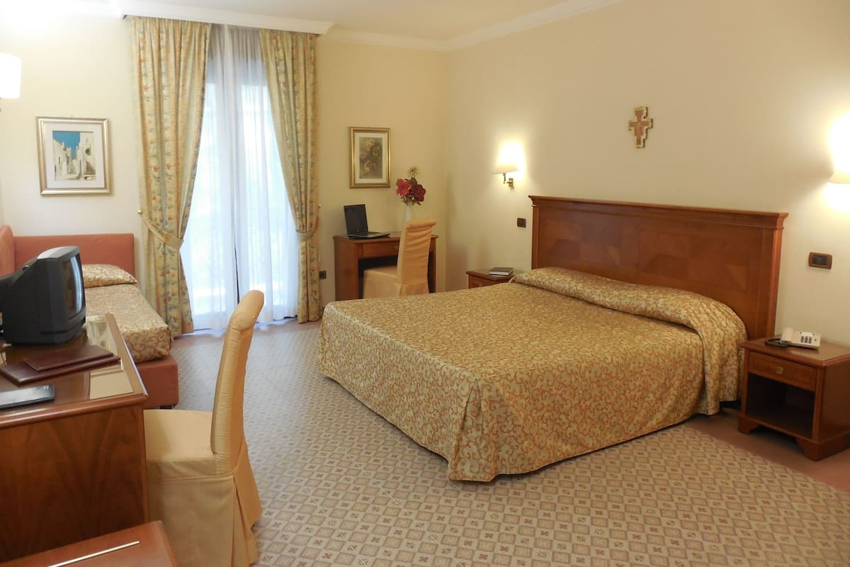 Quarto de hotel com uma cama grande, uma escrivaninha com computador, uma TV e uma cadeira.