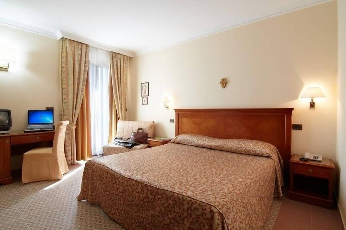 Quarto de hotel com uma cama grande, uma escrivaninha com computador, uma cadeira e uma mesa de cabeceira com abajur.