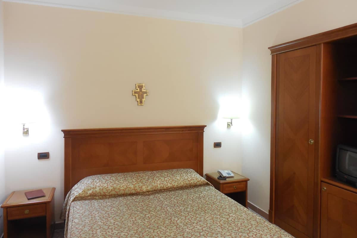 Quarto de hotel com uma cama, um guarda-roupa de madeira, uma mesa de cabeceira com telefone e uma cruz na parede.