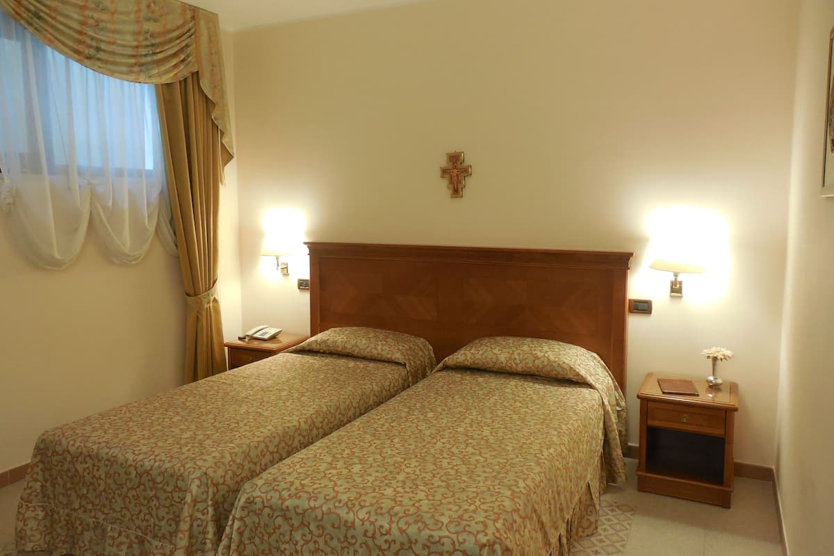 Quarto de hotel com duas camas de solteiro, cabeceira de madeira, luminárias laterais, mesa de cabeceira com abajur, uma cruz na parede e janela com cortinas.