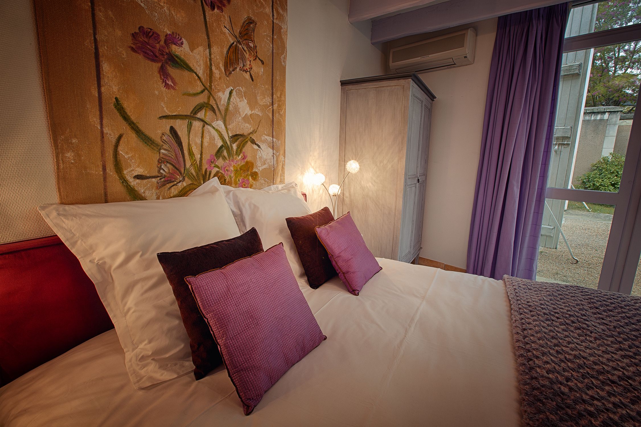 Cama com lençóis brancos e almofadas roxas, tapeçaria com desenhos florais e janela com cortinas roxas.