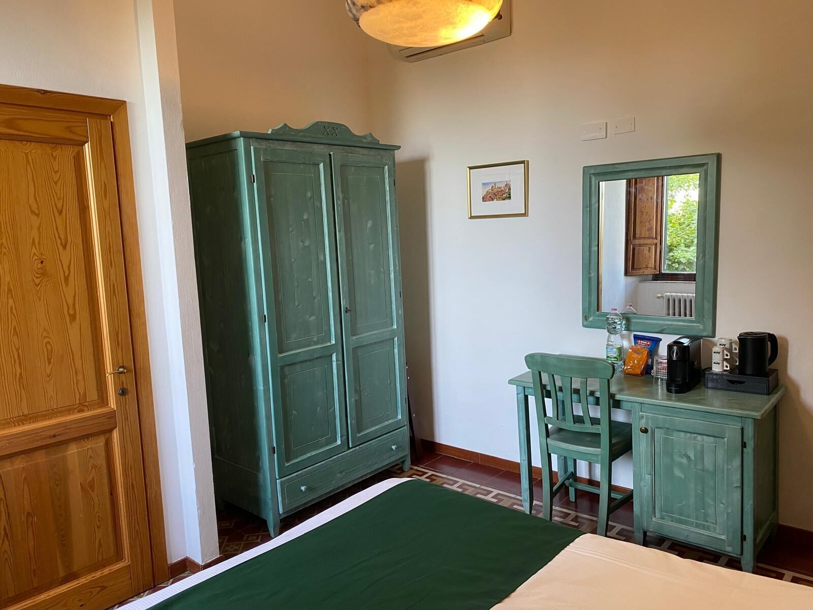 Quarto com porta de madeira, um armário verde, uma escrivaninha com cadeira, um espelho e uma cama com colcha verde.