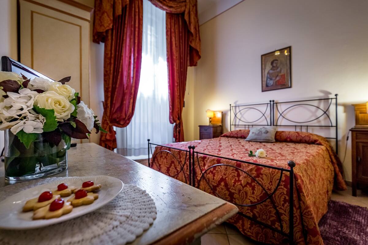 Quarto bem arrumado com uma cama, uma mesa de cabeceira com um prato de biscoitos, um vaso com flores e um quadro na parede.