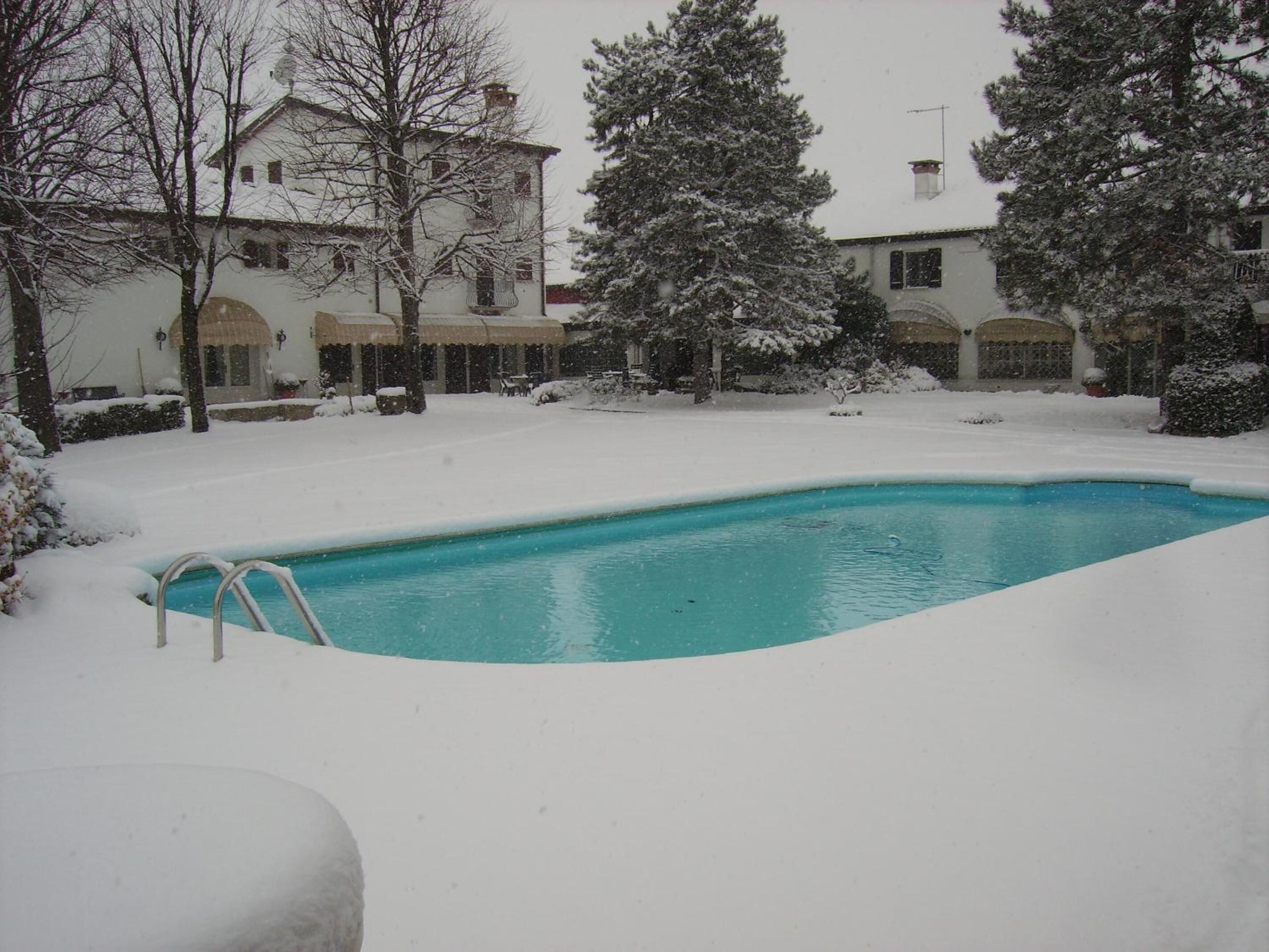 Piscina coberta de neve com superfície de água azul cristalina, rodeada por árvores carregadas de neve e um edifício com várias janelas e uma varanda.