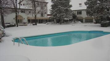 Piscina coberta de neve com superfície de água azul cristalina, rodeada por árvores carregadas de neve e um edifício com várias janelas e uma varanda.