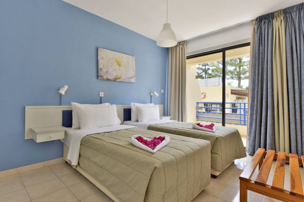 Quarto de hotel com duas camas, mesa de cabeceira, escrivaninha, janela com cortinas e vista para o exterior.