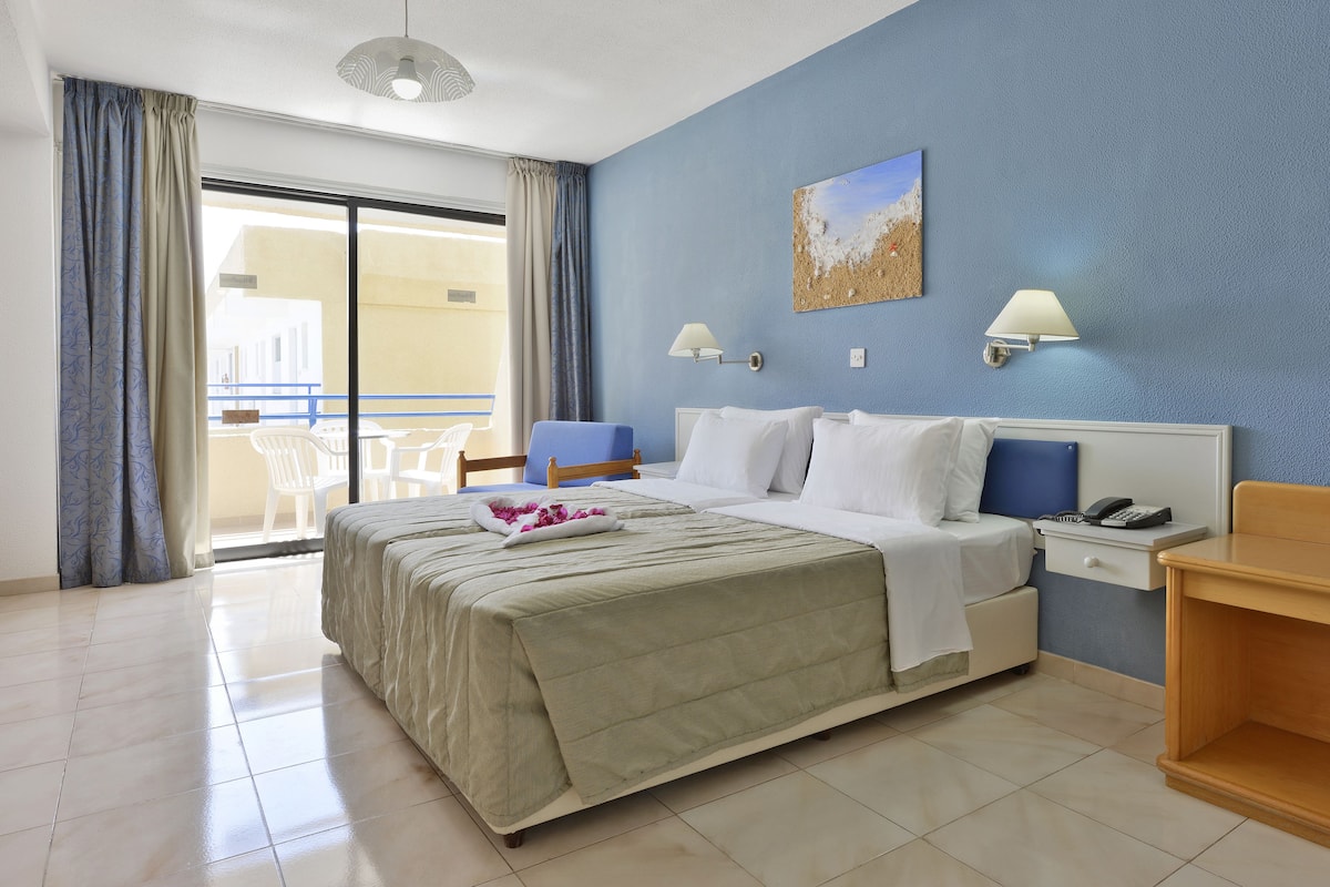 Quarto de hotel com cama, escrivaninha, cadeira, vista para varanda e um quadro na parede.