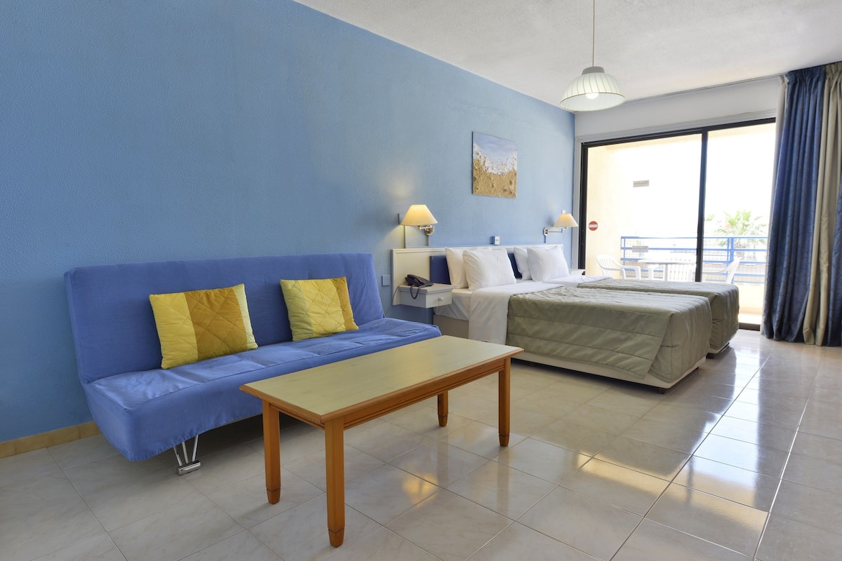 Quarto de hotel com sofá azul, mesa de madeira, cama com colcha branca e varanda com cortinas azuis.