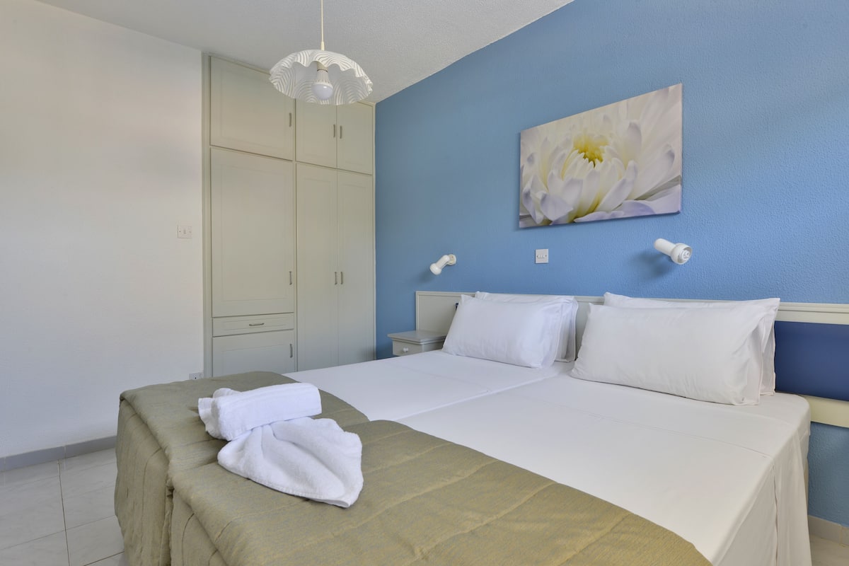 Cama bem arrumada com lençóis brancos e toalha dobrada, mesa de cabeceira branca com abajur, e pintura de uma flor pendurada na parede.