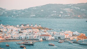 Uma vila costeira com edifícios brancos, um porto com barcos e um cenário de colinas ao fundo.