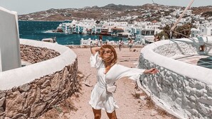 Uma mulher de vestido branco parada em um caminho de terra com paredes de pedra, apreciando a vista de uma cidade costeira com edifícios brancos e barcos na água.