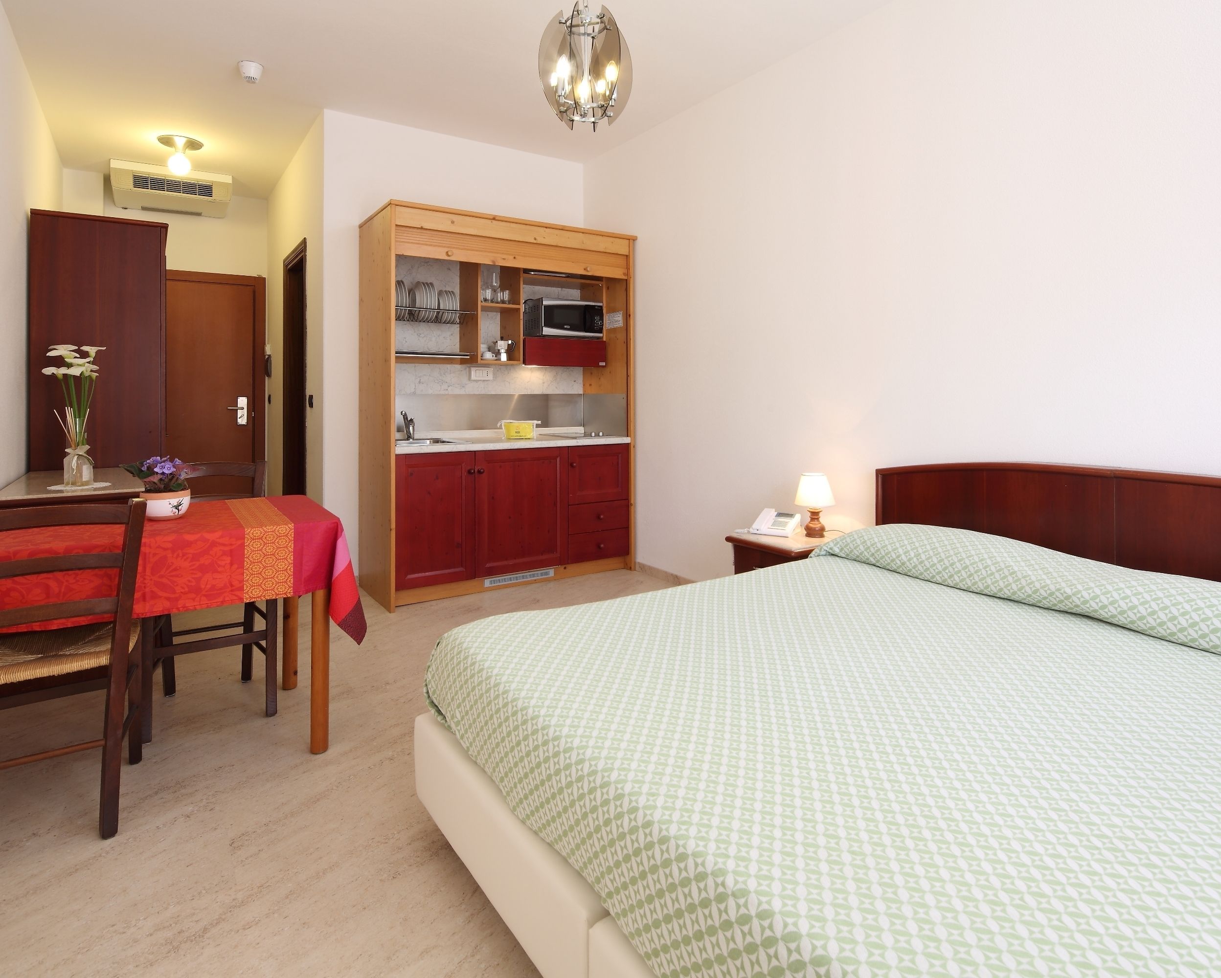Quarto de hotel com cama, secretária, uma cadeira, cozinha compacta e mesa de cabeceira com abajur.