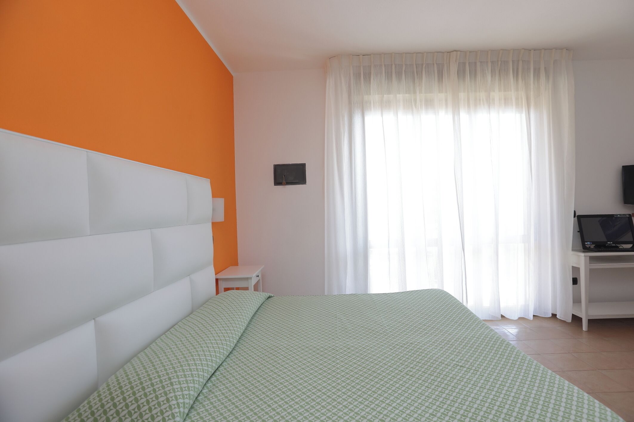 Quarto com cama, uma pequena mesa branca, televisão em um suporte e uma janela com cortinas.