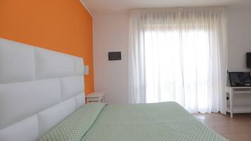 Quarto com cama, uma pequena mesa branca, televisão em um suporte e uma janela com cortinas.