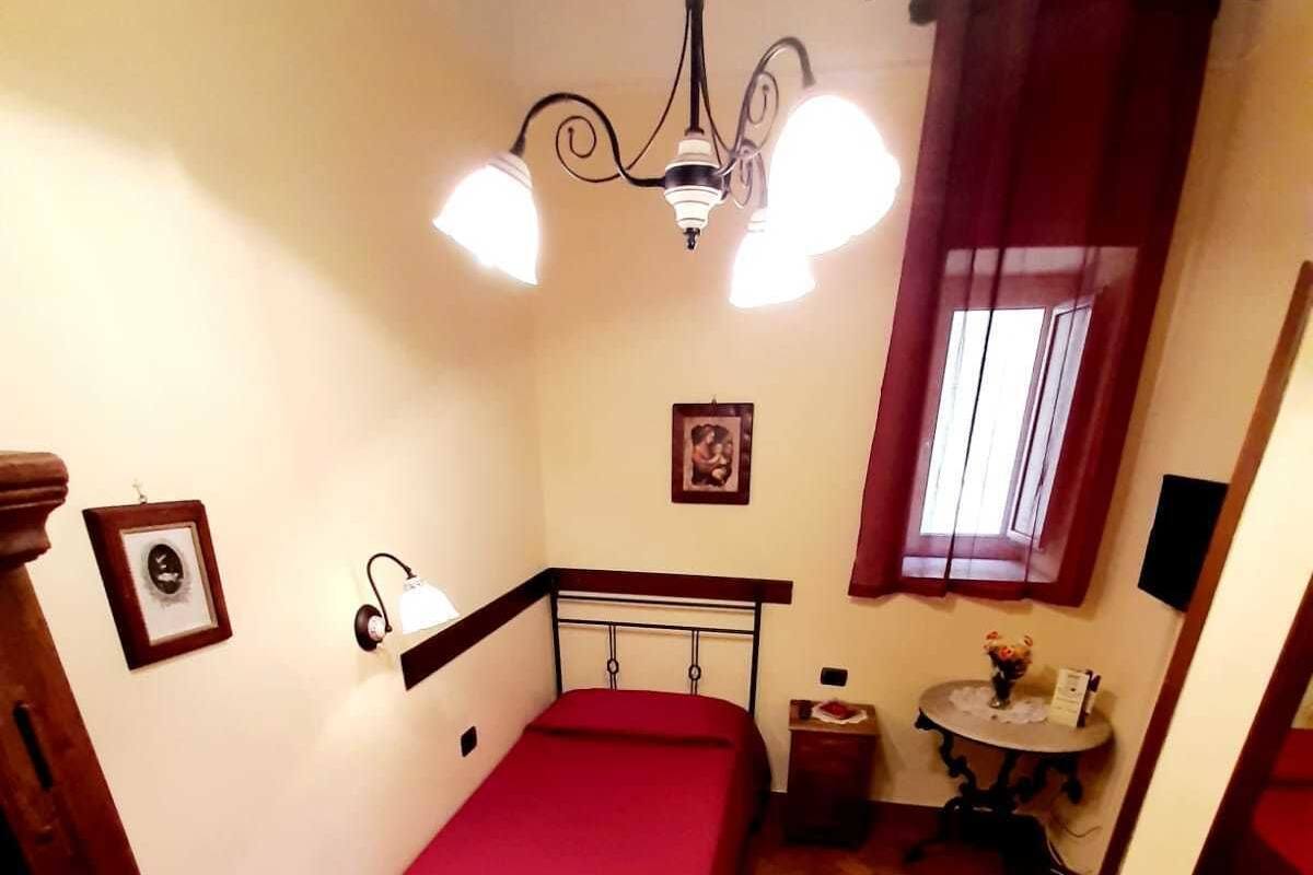 Quarto pequeno e acolhedor com uma cama de solteiro, uma escrivaninha de madeira, uma mesinha, uma cadeira, uma janela com cortinas e um candeeiro de teto.