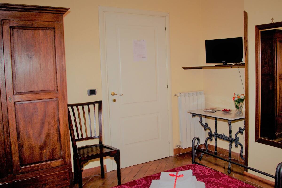 Quarto com guarda-roupa de madeira, porta branca, televisão em uma prateleira, uma mesa pequena com um vaso e uma cadeira.