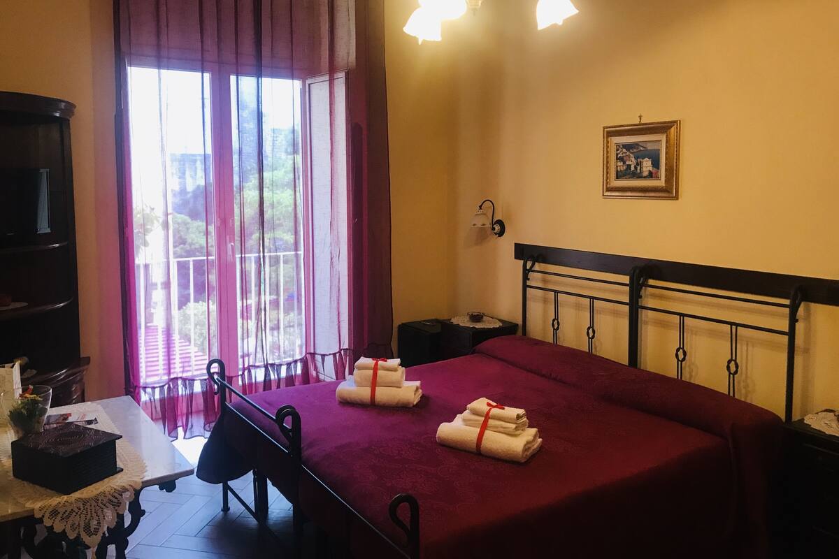 Quarto com cama, mesa de cabeceira, luminária, janela com cortinas, quadro na parede e vista para o exterior.