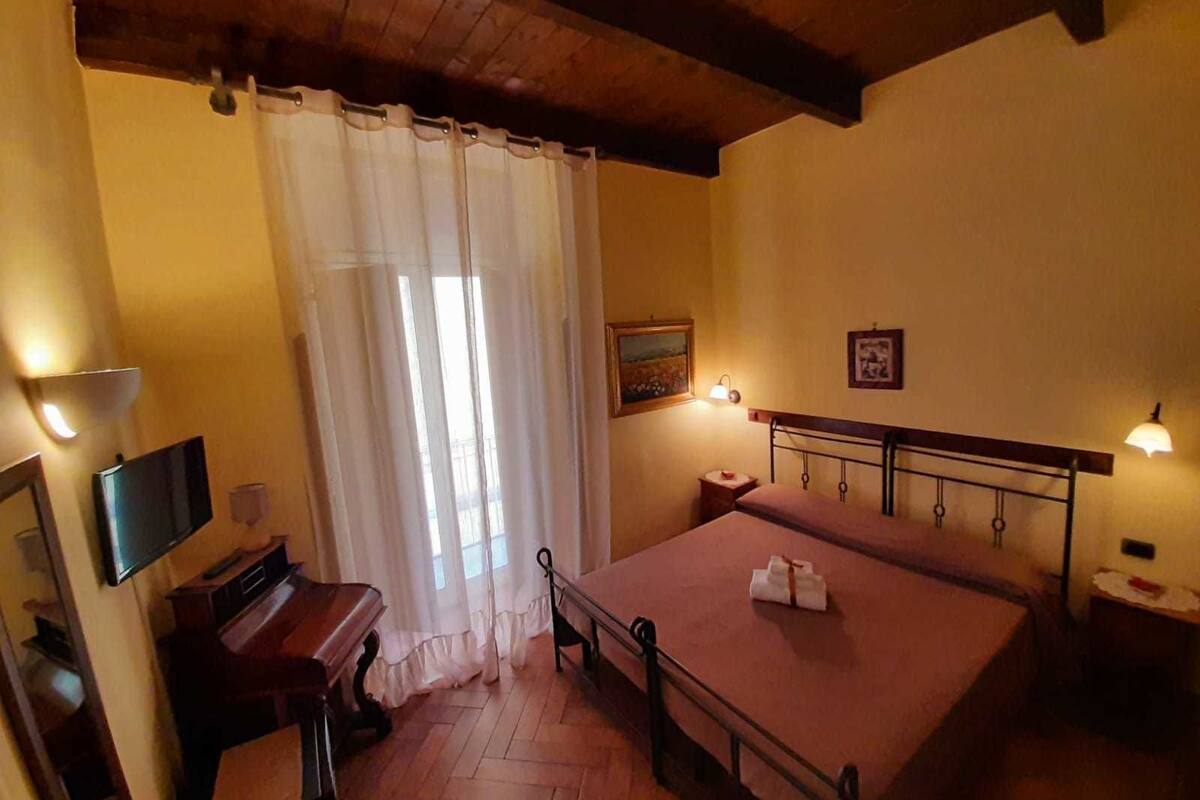 Quarto com cama de madeira, escrivaninha com cadeira, televisão e janela com cortinas leves.