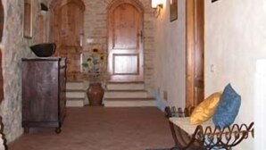 Interior rústico com paredes de pedra, teto de madeira, área de refeições com mesa e cadeiras, e escada que leva a um andar superior.