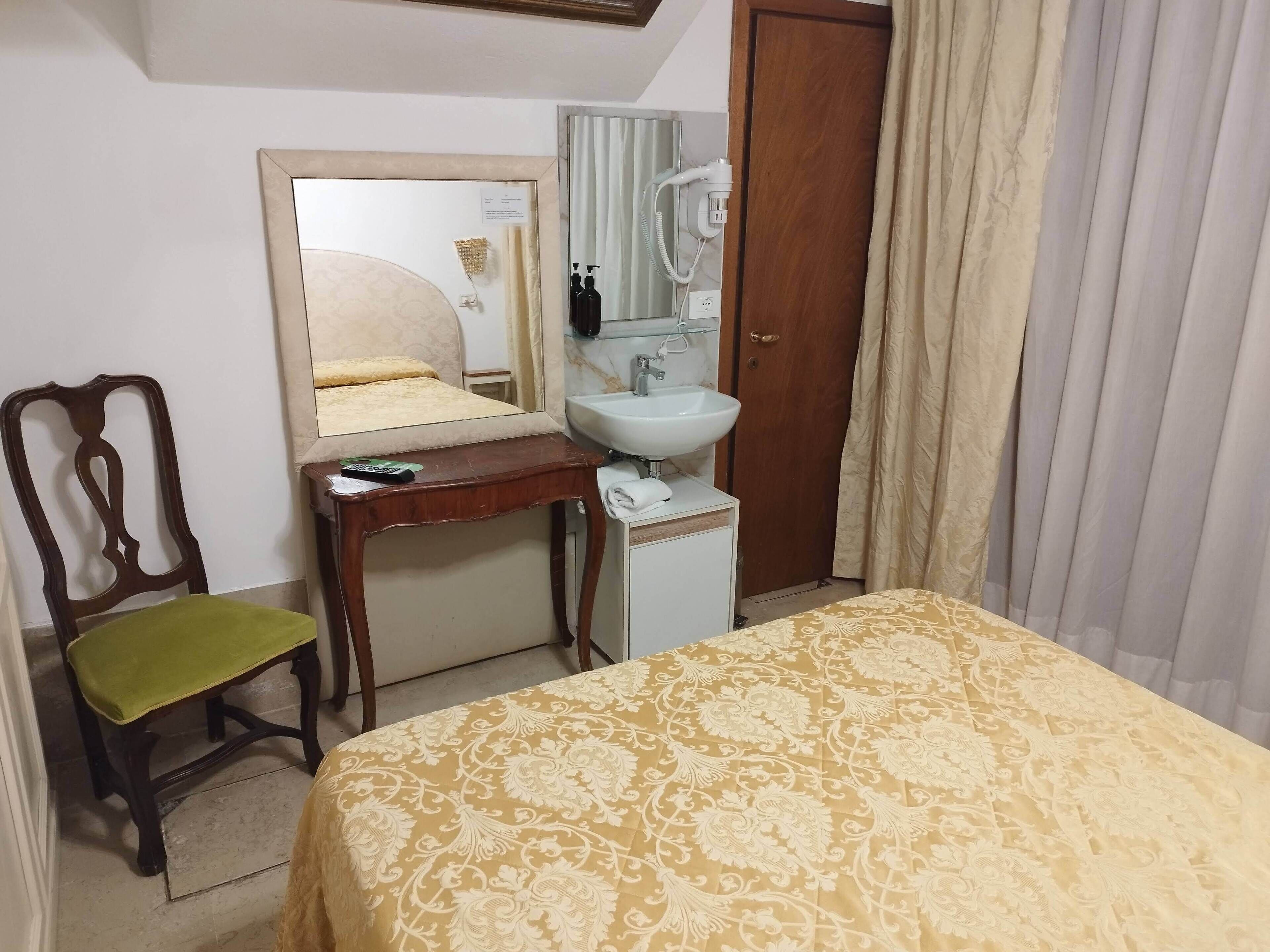 Um quarto pequeno e bem organizado, com uma cama de solteiro, uma cadeira de madeira, uma mesinha, um espelho e um banheiro com pia e secador de cabelo.