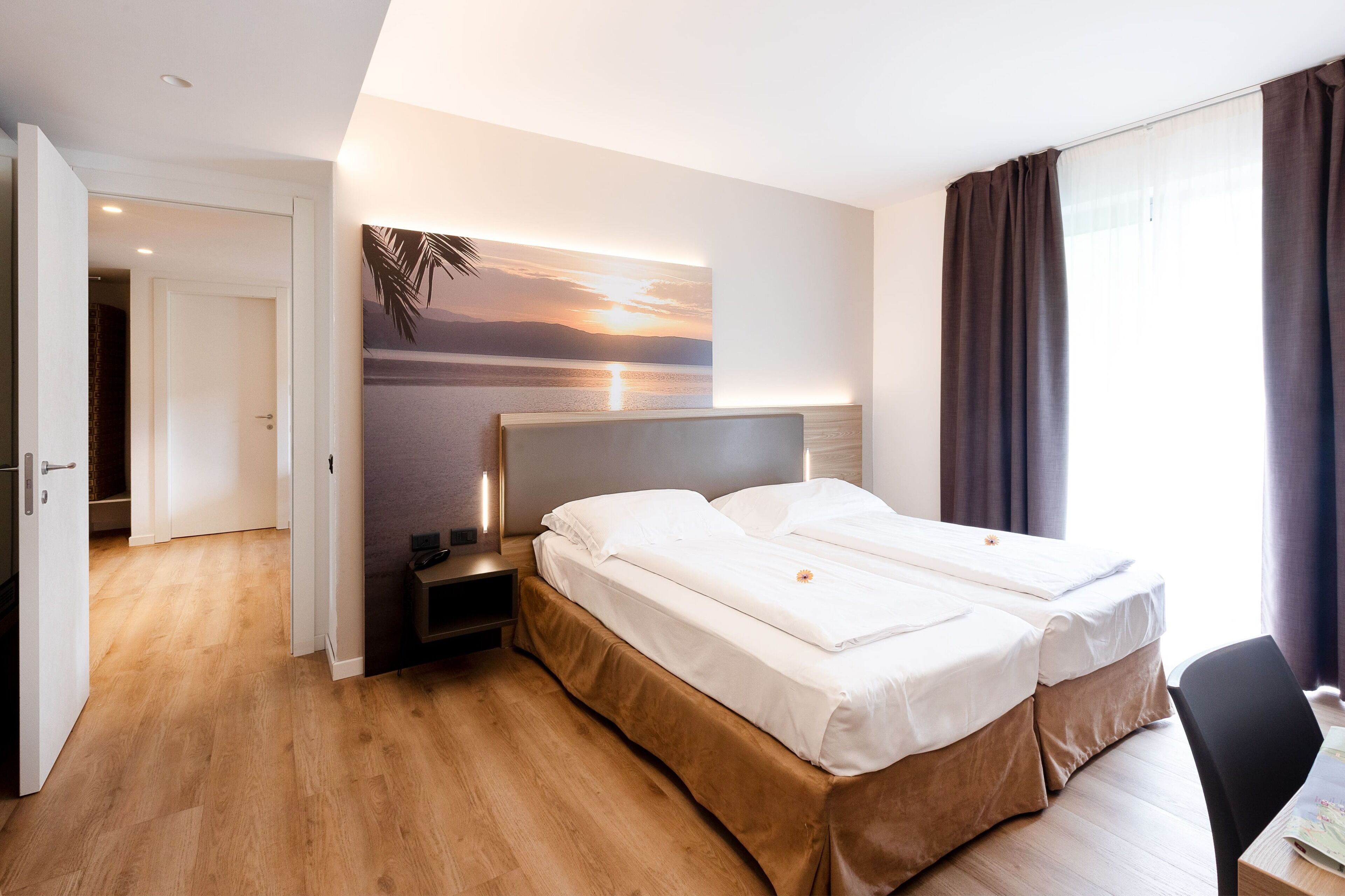 Quarto de hotel com uma cama grande, mesa de cabeceira, escrivaninha e vista para o pôr do sol pela janela.