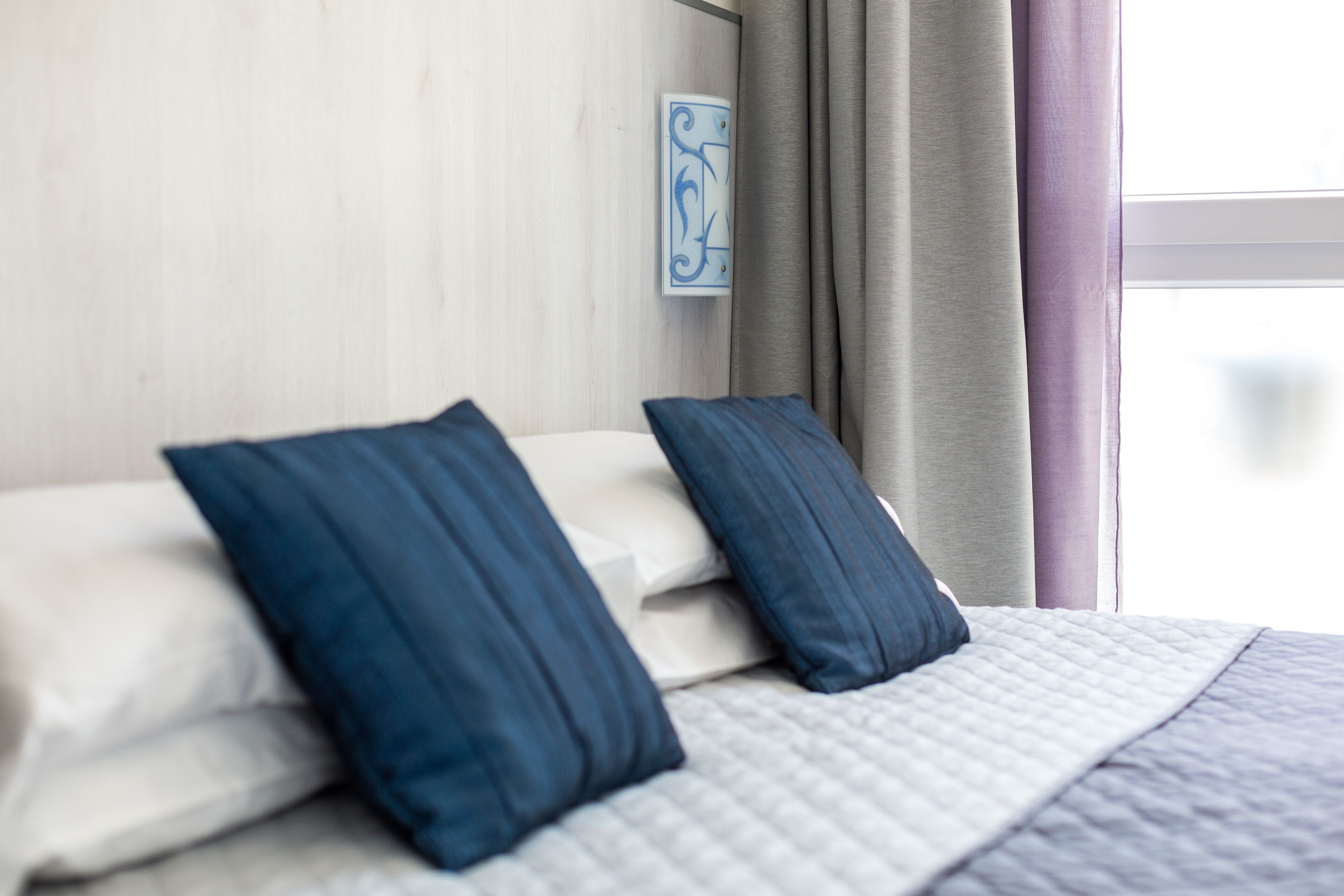 Uma cama com almofadas azuis e brancas, um edredom claro e uma janela com cortinas.
