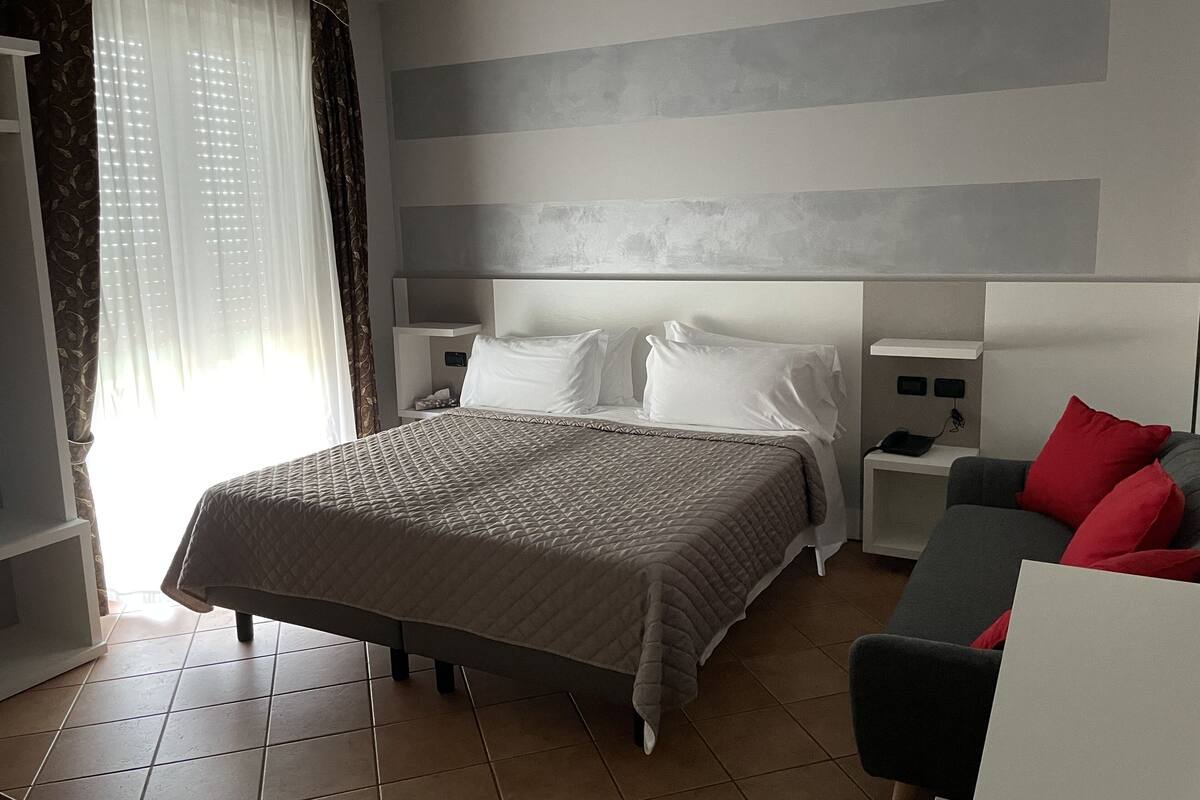 Quarto de hotel com uma cama grande, duas mesinhas de cabeceira, um criado-mudo, um sofá cinza com almofadas vermelhas e uma janela com cortinas finas.