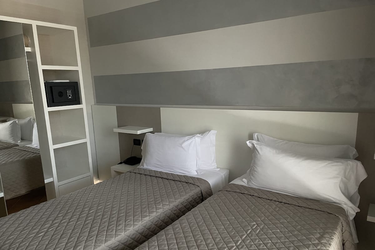 Quarto de hotel com duas camas, cada uma com colcha cinza acolchoada e almofadas brancas. Há uma mesa de cabeceira branca com um telefone preto e uma pequena prateleira.