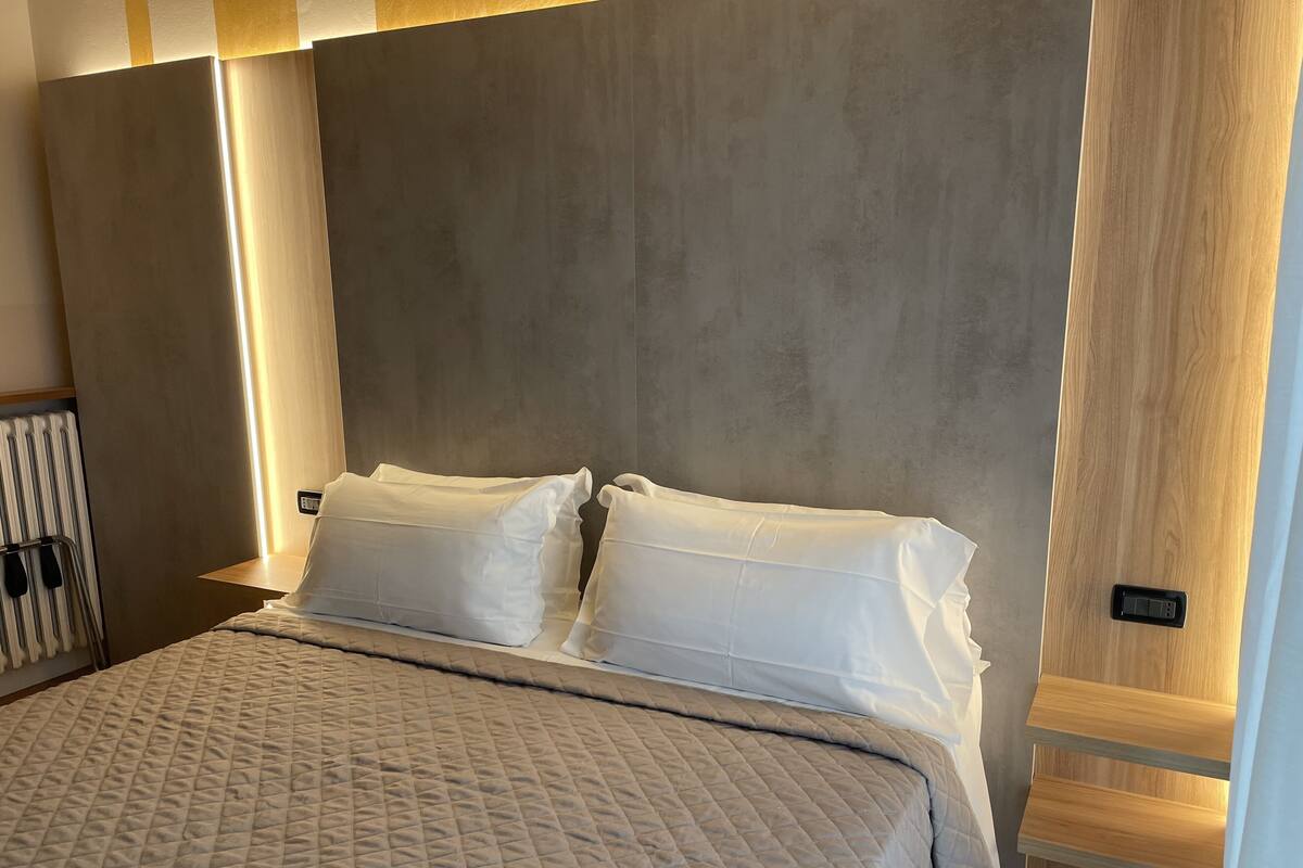Quarto moderno com uma cama grande, cabeceira com iluminação embutida e radiador à esquerda.