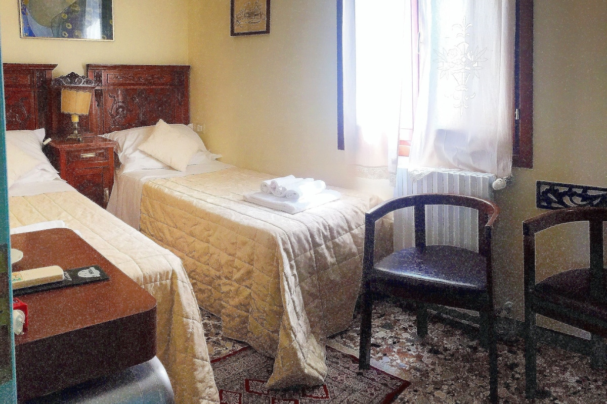 Quarto de hotel com duas camas, uma mesa de cabeceira de madeira, uma cadeira, uma escrivaninha e uma janela com cortinas.