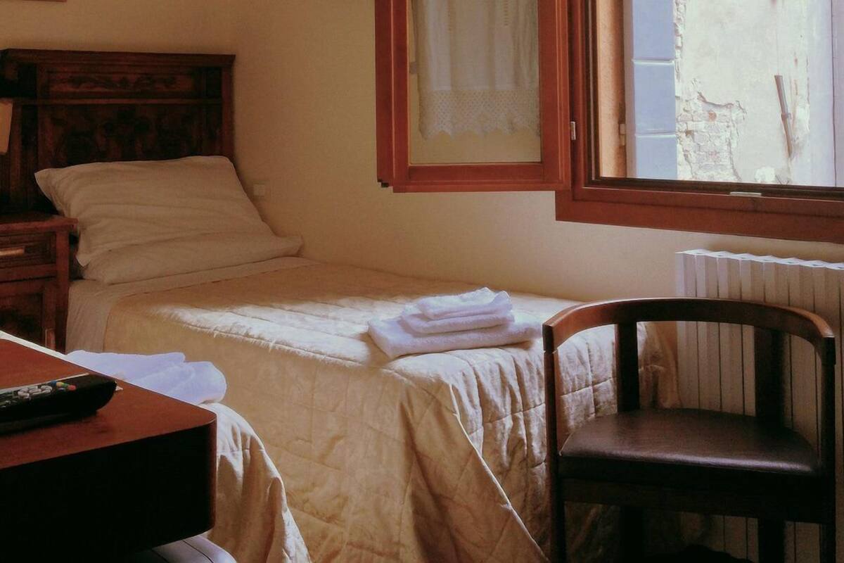 Quarto com cama, cadeira, mesa de cabeceira e janela com vista para uma parede de tijolos.