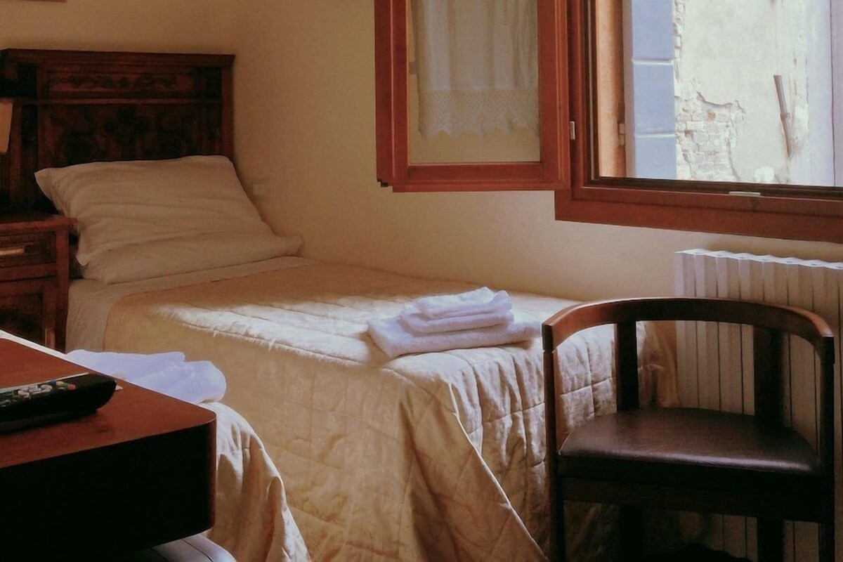 Uma cama de solteiro com cabeceira de madeira, mesa de cabeceira com controle remoto, uma cadeira e uma janela com vista para um prédio.