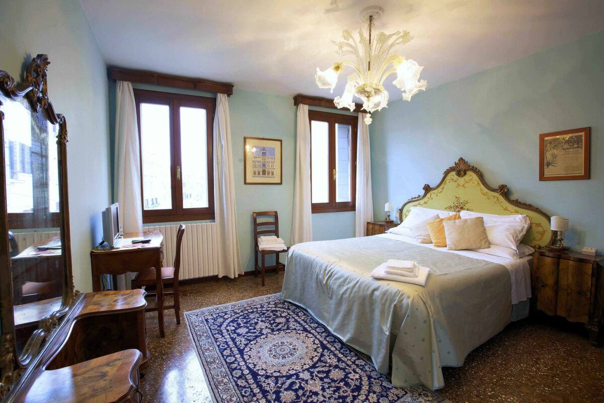 Quarto com cama grande, escrivaninha, cadeira, candelabro e janela com cortinas.