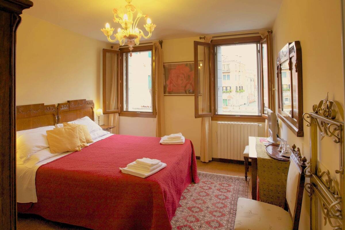 Quarto de hotel com uma cama grande, um candelabro, uma janela com vista para edifícios, um radiador e uma mesa com espelho.