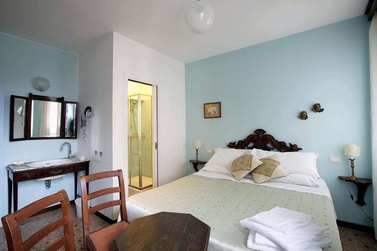 Quarto de hotel com cama, escrivaninha, cadeira, banheiro e chuveiro.