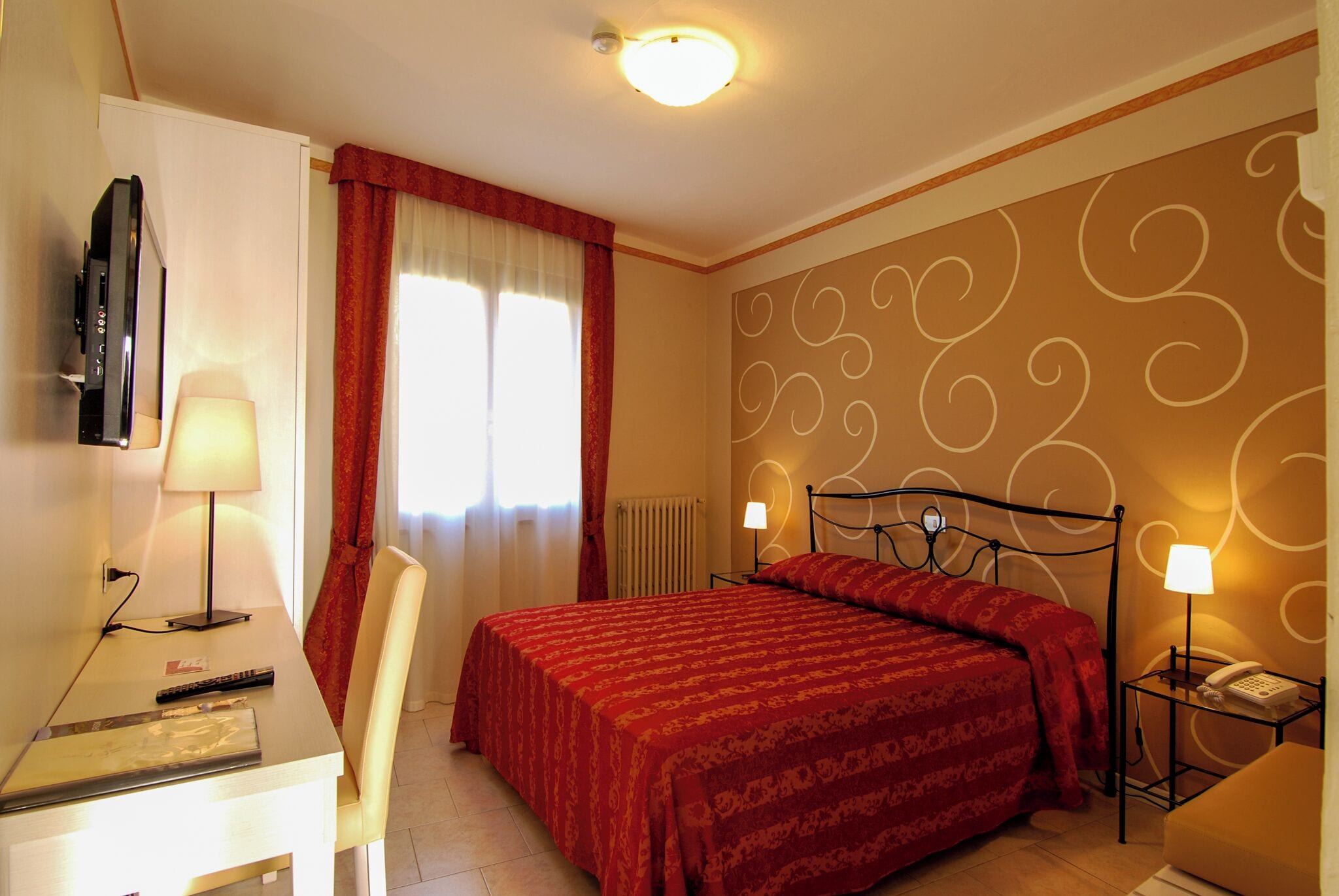 Quarto de hotel com uma cama, uma escrivaninha com luminária, uma cadeira, uma televisão e uma mesa de cabeceira com luminária e telefone.