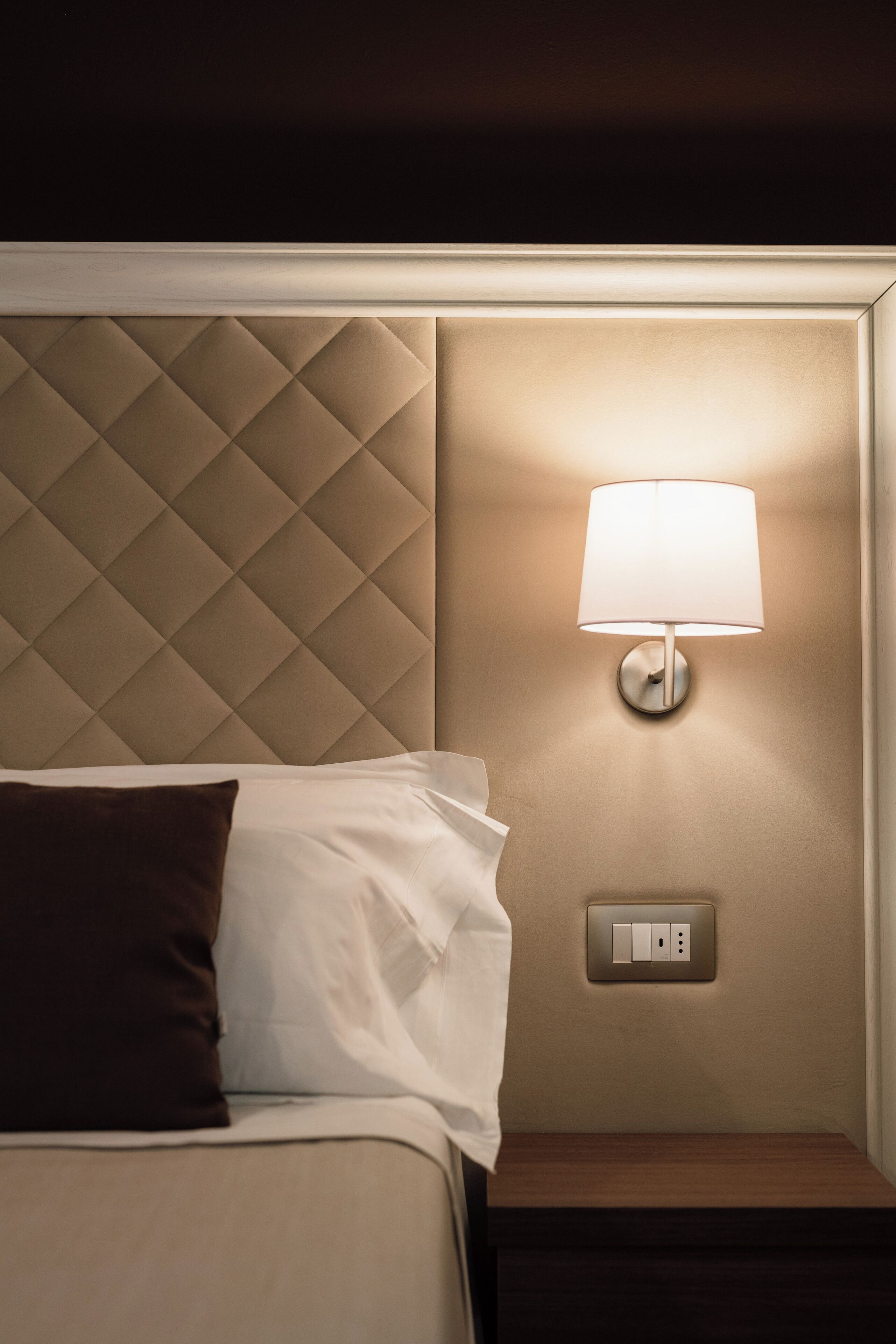 Quarto de hotel com uma cama, mesa de cabeceira, luminária na parede e tomada.