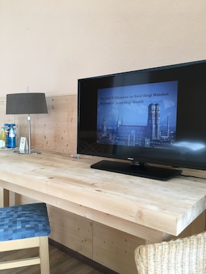Televisão de tela plana em uma mesa de madeira com uma vista urbana ao fundo, uma luminária e água engarrafada.