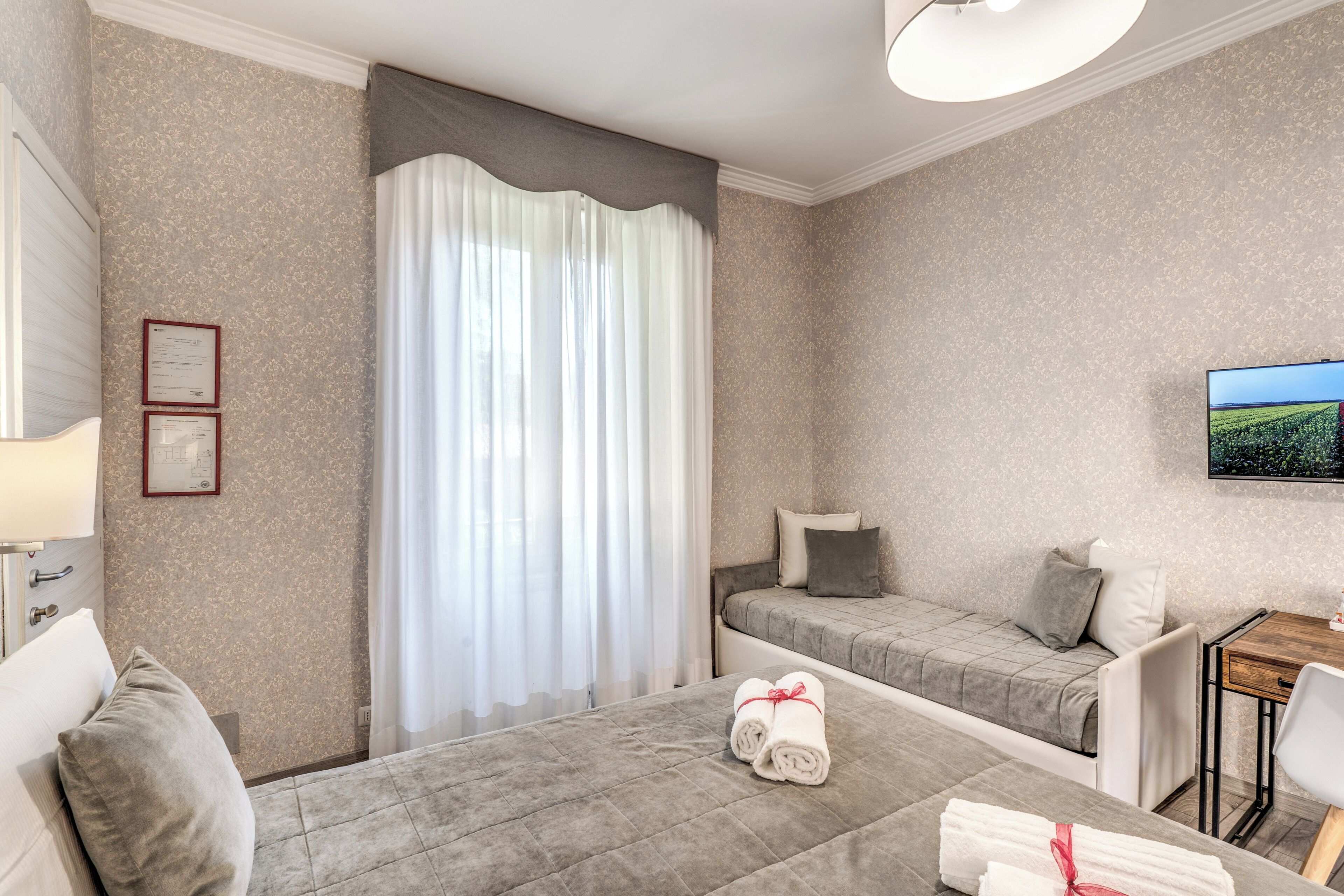 Quarto de hotel com uma cama grande, duas camas menores, uma escrivaninha, uma cadeira, uma televisão e uma janela com cortinas.